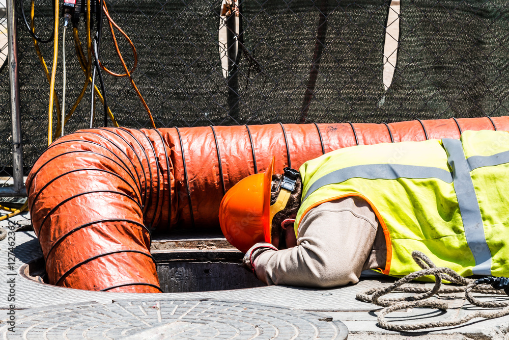 Worker wearing safety gear and looking down manhole in an urban setting ...
