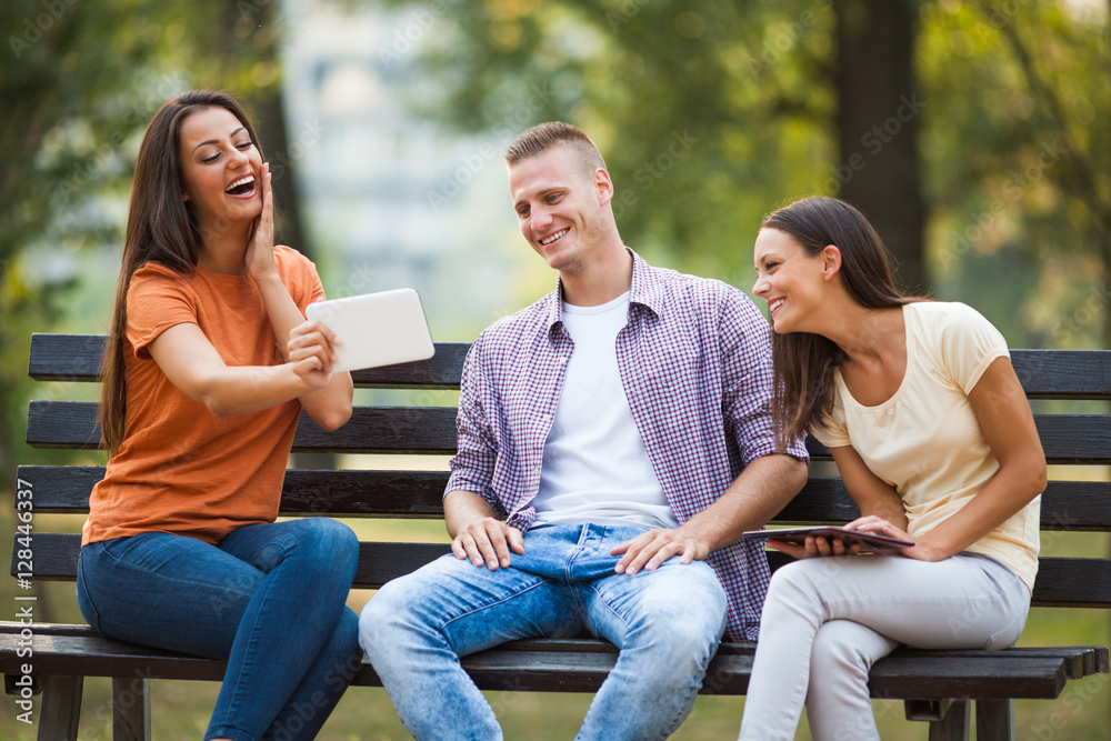 Three friends are sitting on bench in park and talking. They are using ...