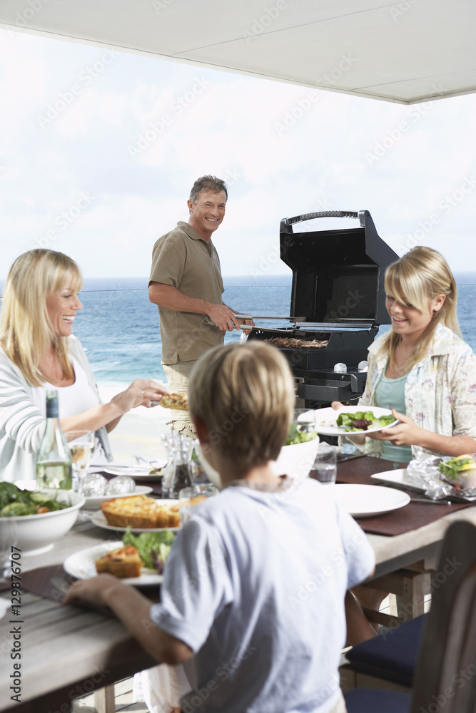 Family sitting around table eating barbecued food with father standing ...