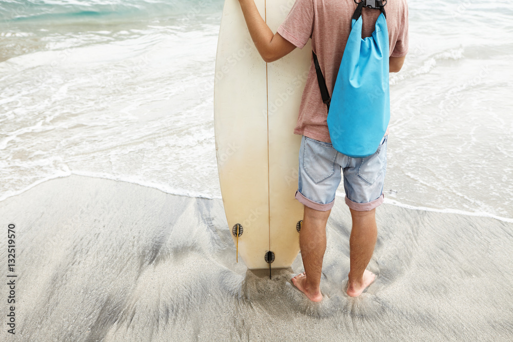 Rear portrait of surfer standing barefooted on beach in sea water ...