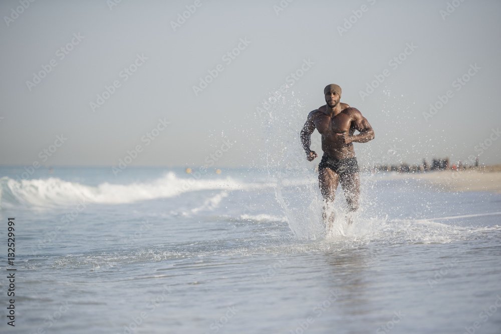 Dark muscular man running on water edge of the beach splashing water ...