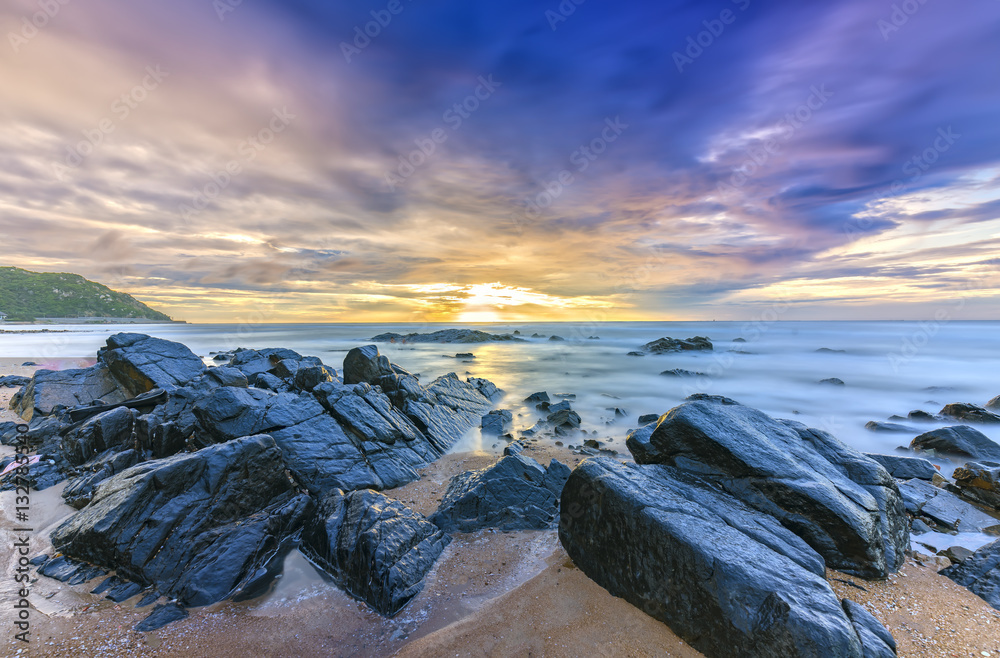 Dawn on the beach with brightly colored clouds, beneath a rock with ...