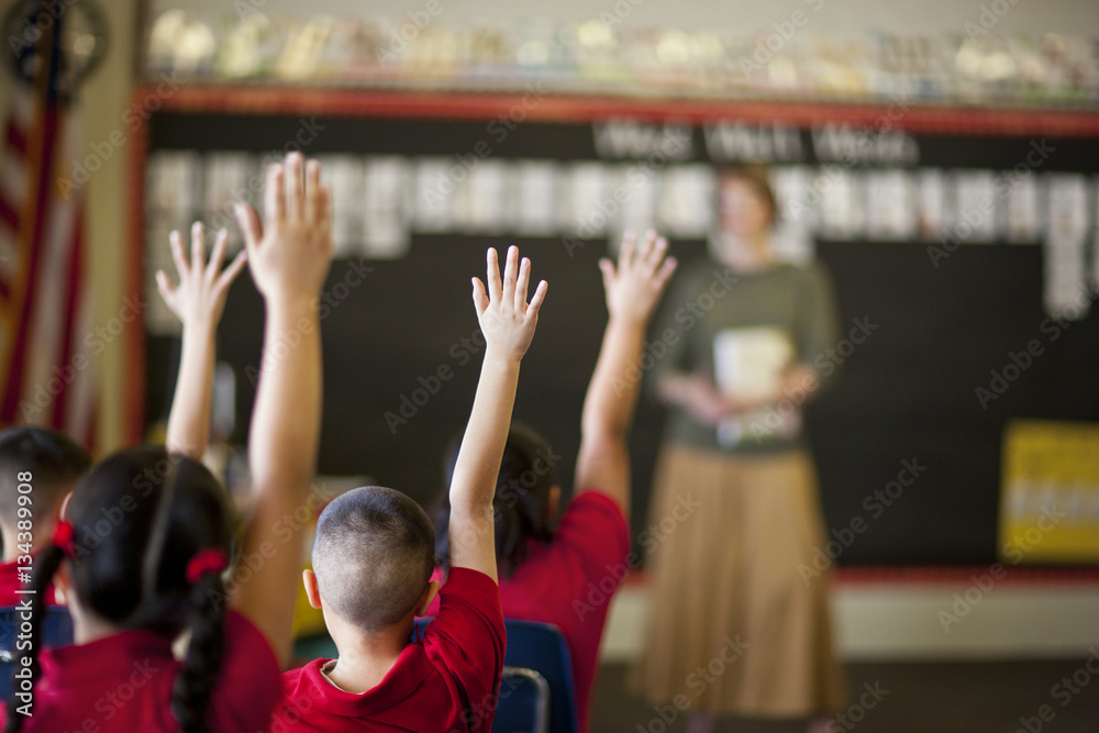 Students with their hands raised in a classroom. Stock Photo | Adobe Stock