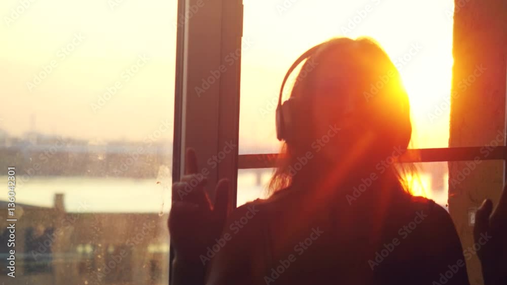 Young daydreaming woman sitting by the window wears headphones through ...