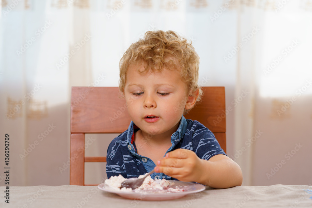 Blond curly-haired boy lefty eats cottage cheese for breakfast, sitting ...