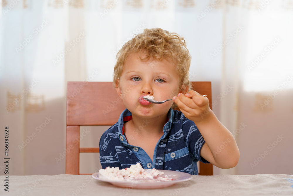 Blond curly-haired boy lefty eats cottage cheese for breakfast, sitting ...