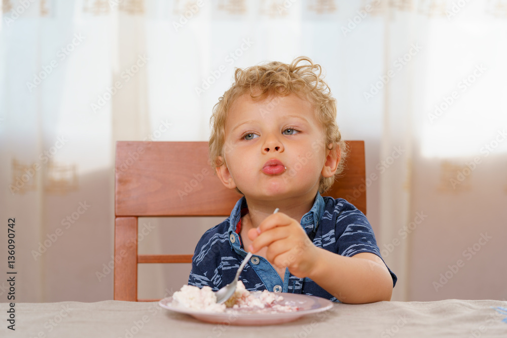 Blond curly-haired boy lefty eats cottage cheese for breakfast, sitting ...