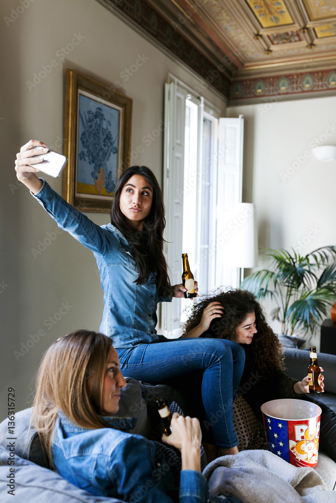 Young woman with beer bottle taking a selfie while seating on the couch ...