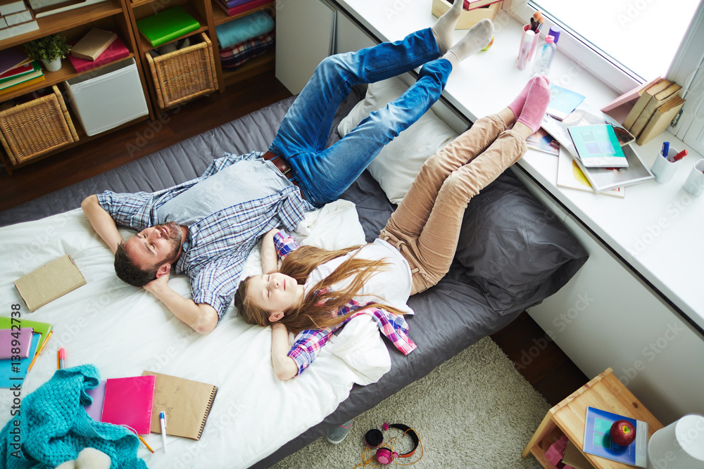 Father and his teenage daughter lying on bed with legs on window sill ...