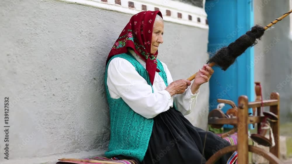 Elderly woman sits on bench and works with distaff with spinning wheel ...