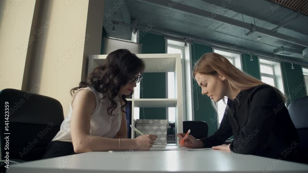 Two business women in office discuss topics, watching documents. blonde ...
