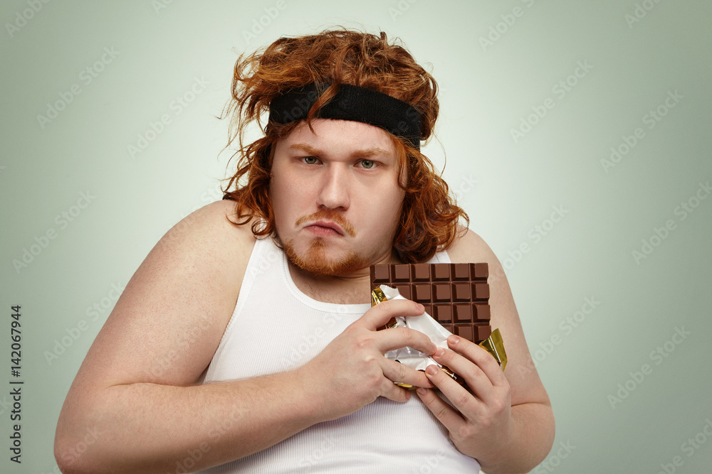 Greedy obese fat young man wearing sports band on curly ginger hair ...
