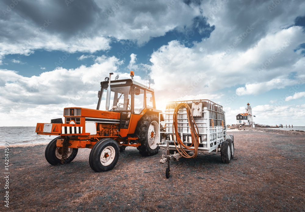 Red tractor with trailer against lighthouse and blue overcast sky in ...
