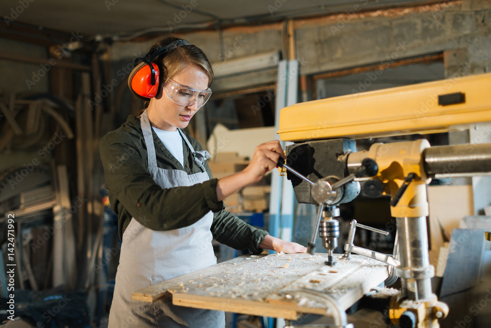Pretty young carpenter wearing ear and eye protectors while using drill ...