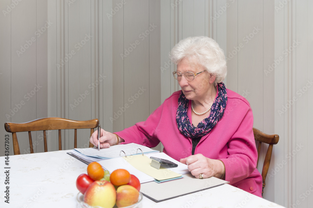 Alte Frau sitzt am Tisch über einem Aktenordner und rechnet etwas aus ...