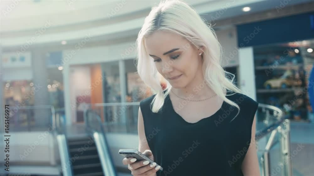 Happy young blonde woman using cell phone at a shopping center ...