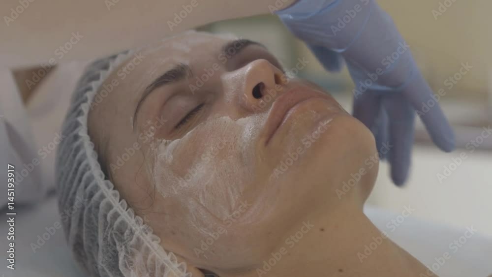 Close-up of a young cosmetologist puting cream on the face of client ...