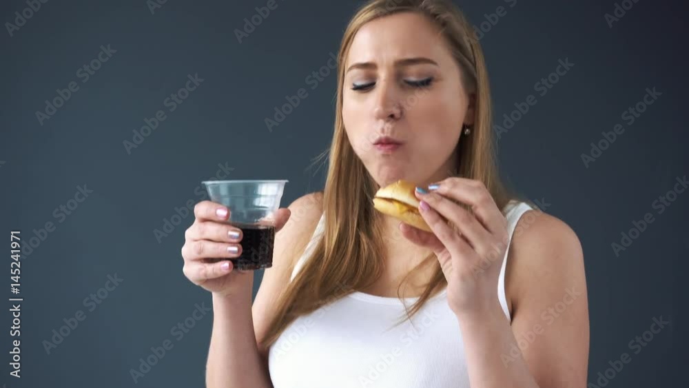 Overweight woman eating a hamburger and drinks a soda on the grey ...