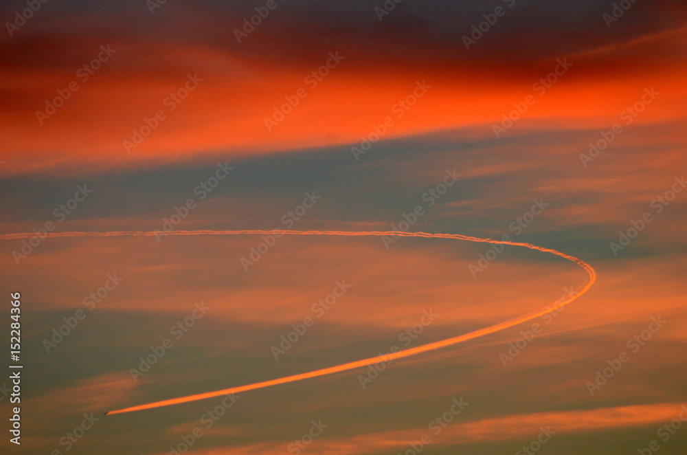 Red contrail on a blue sky at sunset. A plane makes an U turn. There is ...