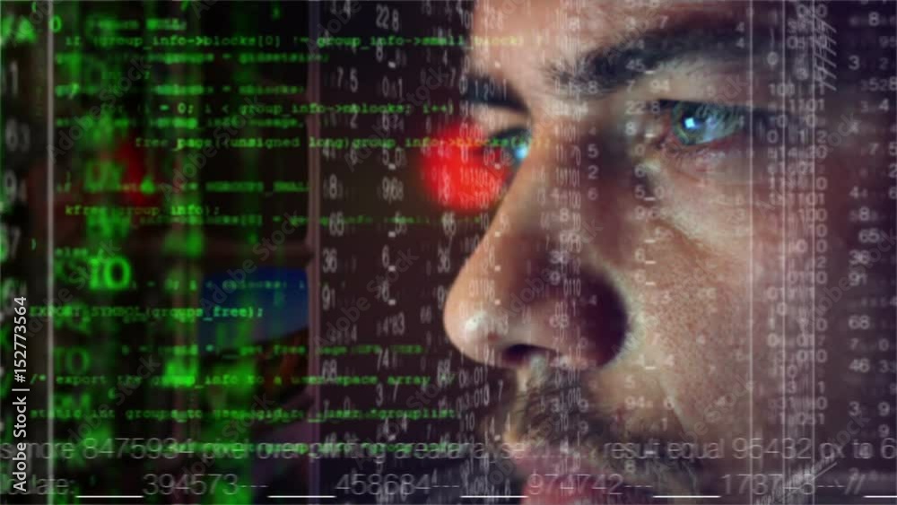 Male hacker working on a computer while green code characters reflect on his face in a dark ...