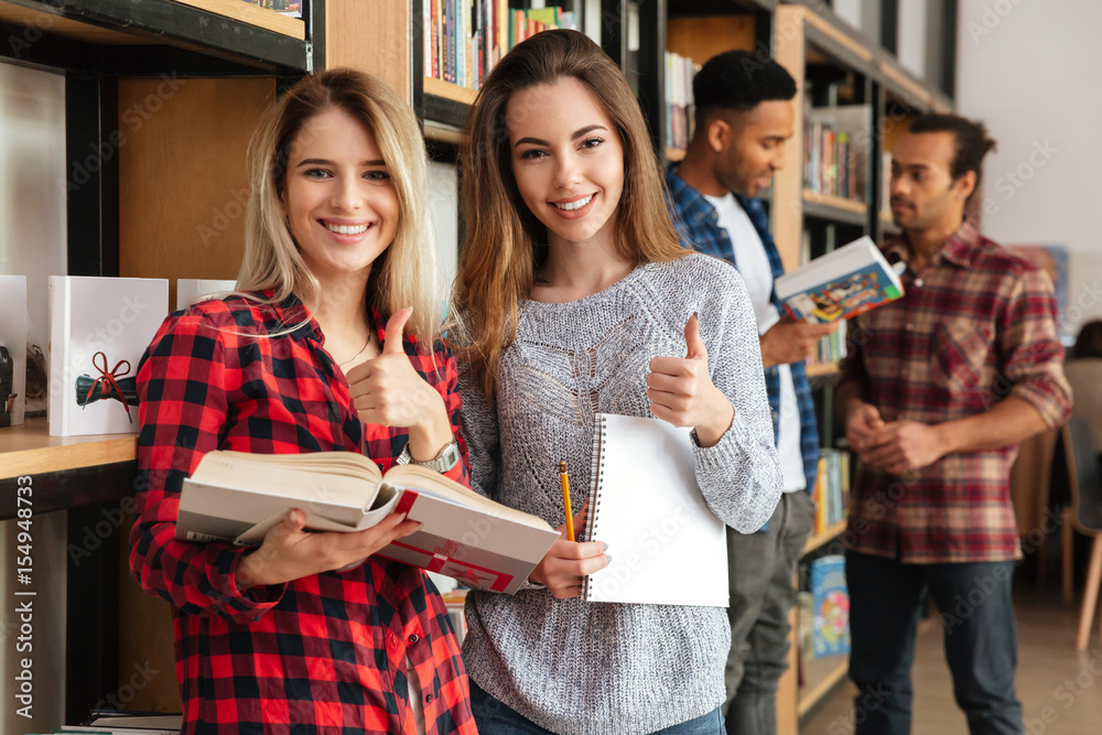 Happy women students standing in library reading books. Stock Photo ...