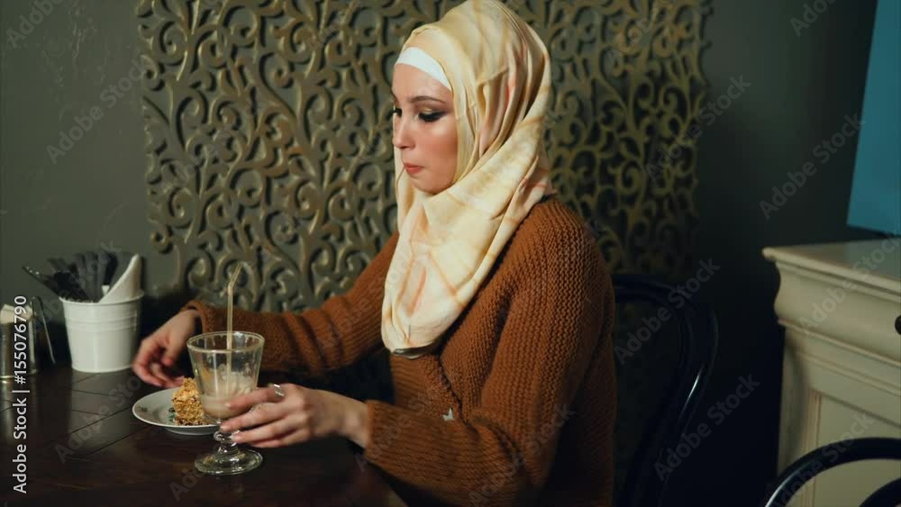 A modern and young Muslim woman in a hijab eats a cake and drinks a ...