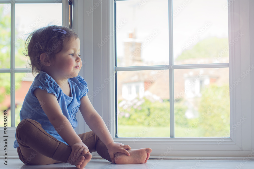 Toned photo of little toddler girl sitting on the window looking ...
