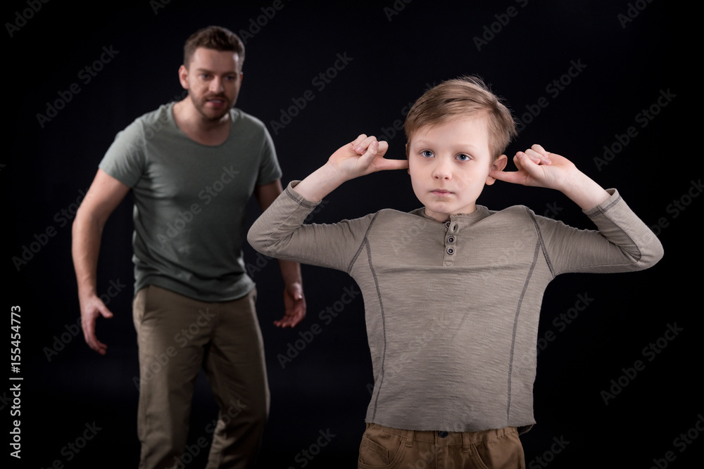 Angry father looking at son standing and closing ears with fingers ...