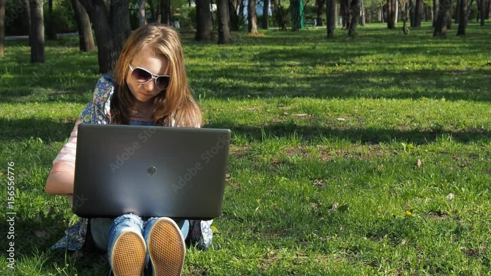 Teenager working on laptop in nature. The girl is sitting on the grass ...