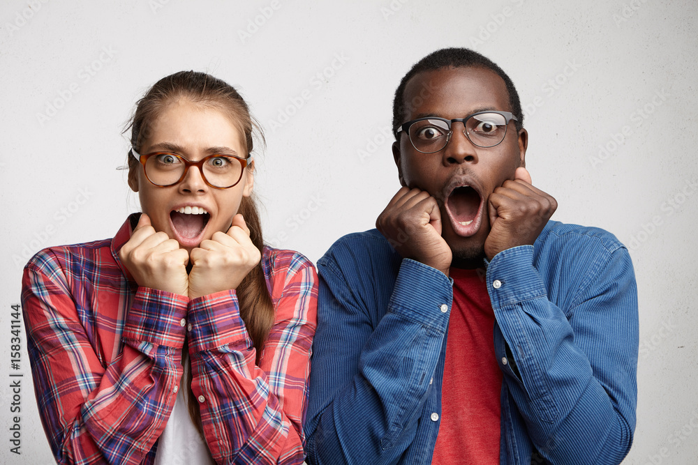 Terrified and puzzled couple with opened mouths holding fists under ...