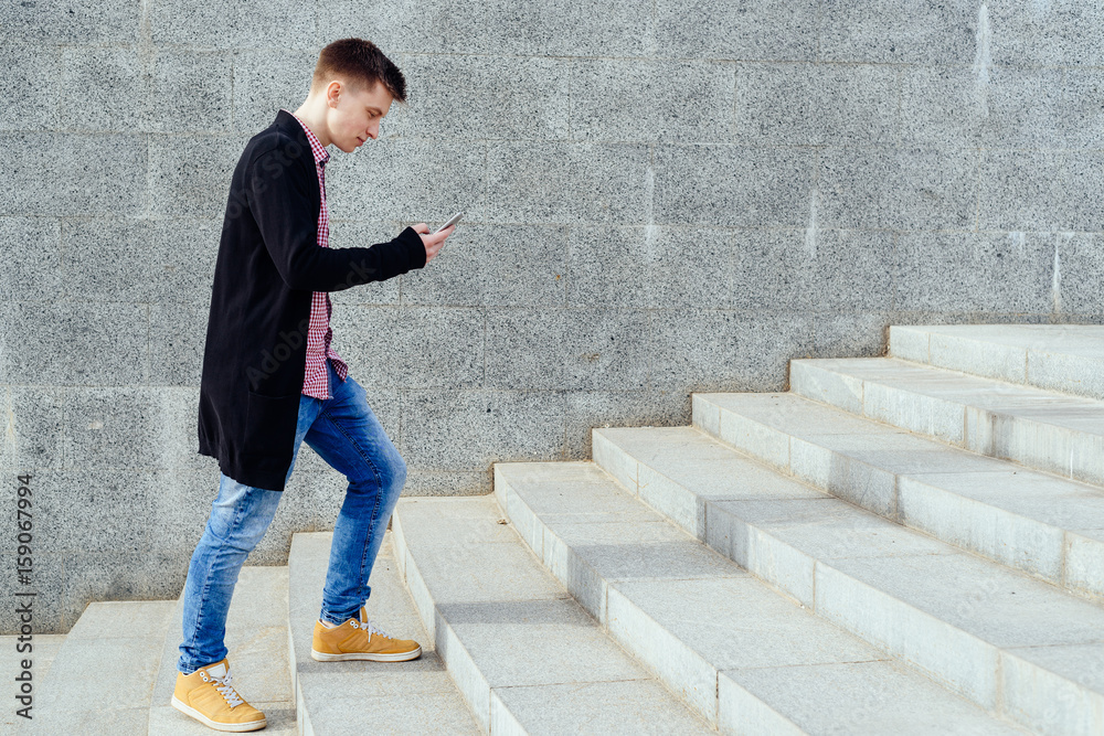 Stylish young man in plaid shirt and jeans walking up stairs and using ...