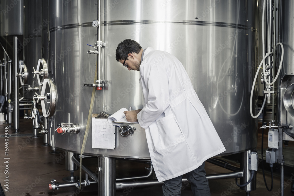 Side view of brewer in brewery wearing lab coat writing on clipboard ...