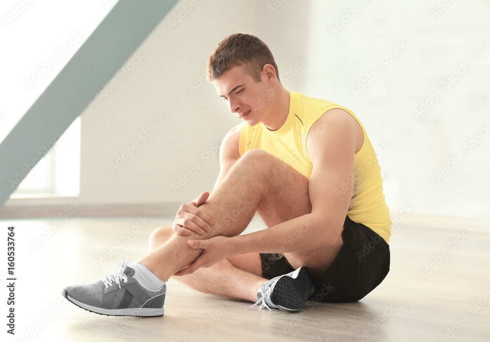Young man suffering from pain in leg while sitting on floor indoors ...