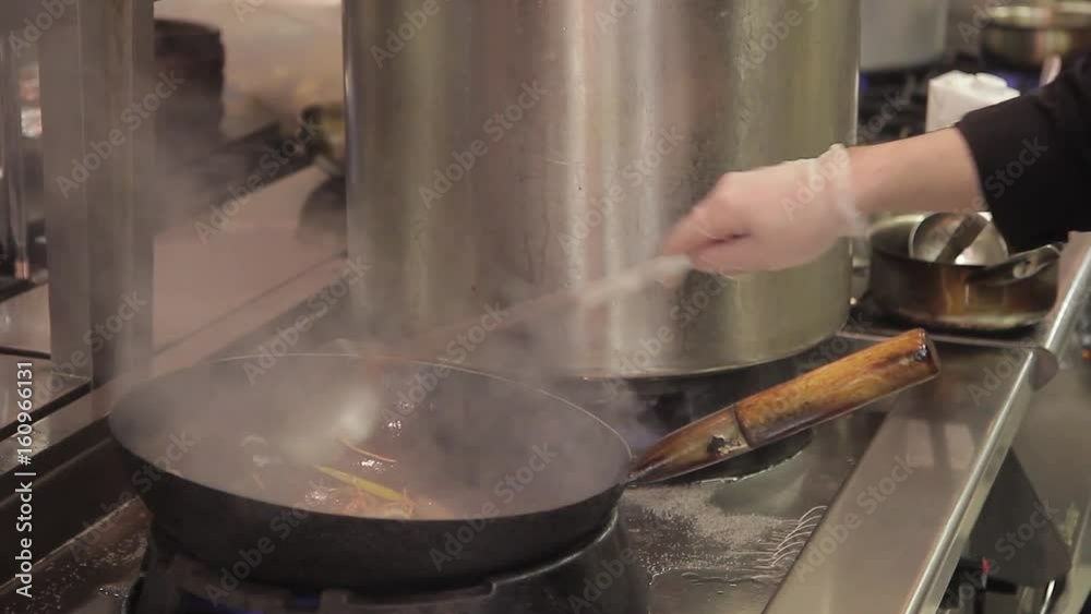 Mixing udon and vegetables in frying-pan at stove in industrial ...