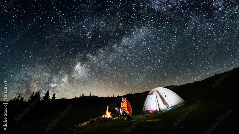 Night camping. Couple tourists sitting at a campfire near illuminated ...