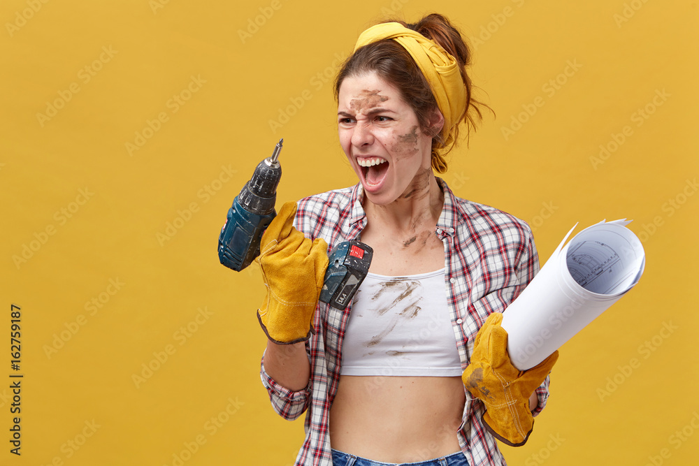 Angry female industrial worker holding rolled paper and drill looking ...