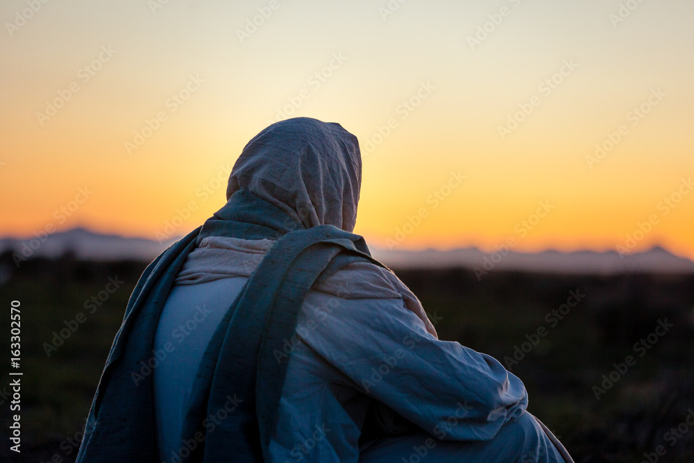 Poster Jesus Christ sits on a hill side and watches the sun set on the ...