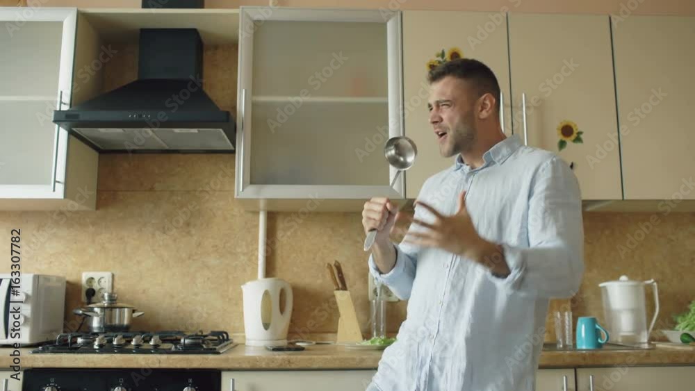 Attractive young funny man dancing and singing with ladle while cooking ...