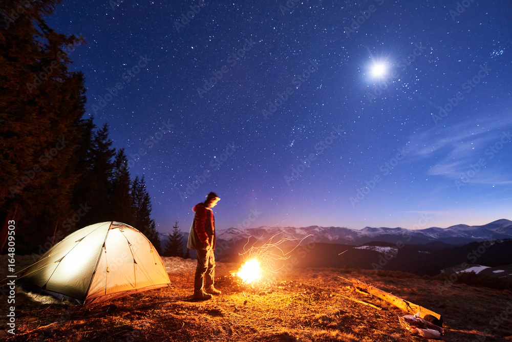 Male tourist have a rest in his camp at night, standing near campfire ...