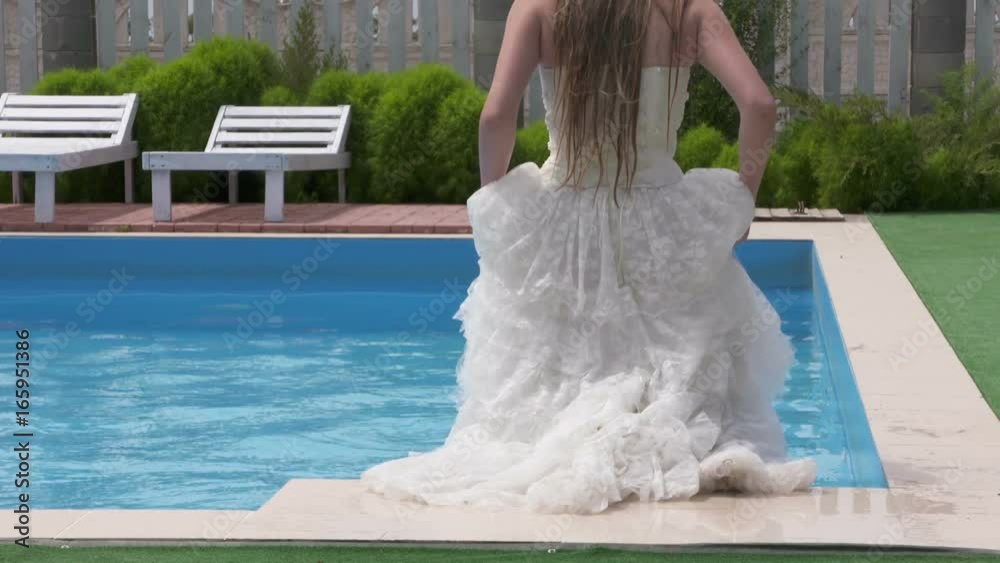 Young girl in wet wedding dress descending the stairs in the pool. Rear ...
