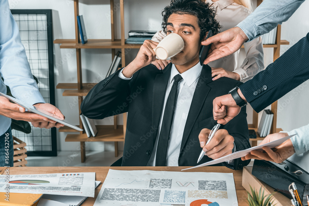 portrait of busy businessman drinking coffee and sitting at workplace while colleagues helping ...