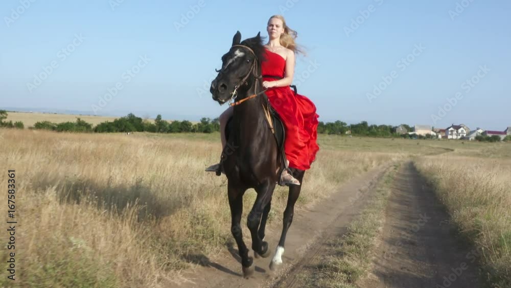 Young girl wearing long red dress riding black horse in countryside ...