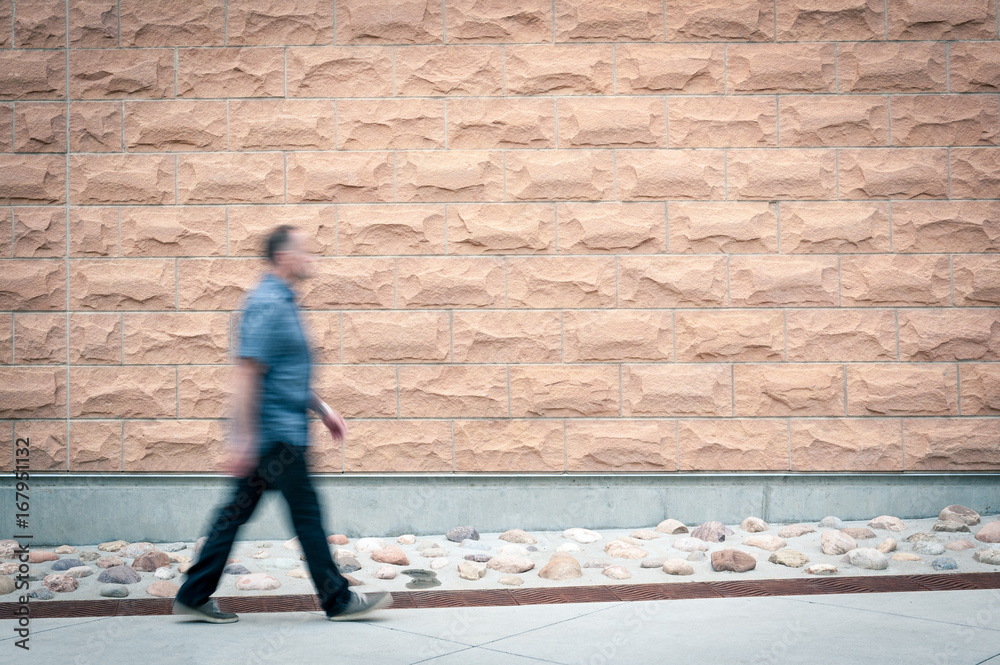 Man walking down sidewalk in front of a large blank brick rock wall ...