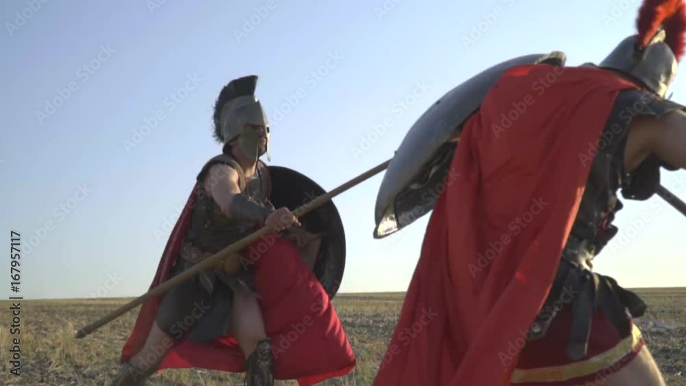 Roman gladiators in heavy armor and red cloaks beat on spears and ...