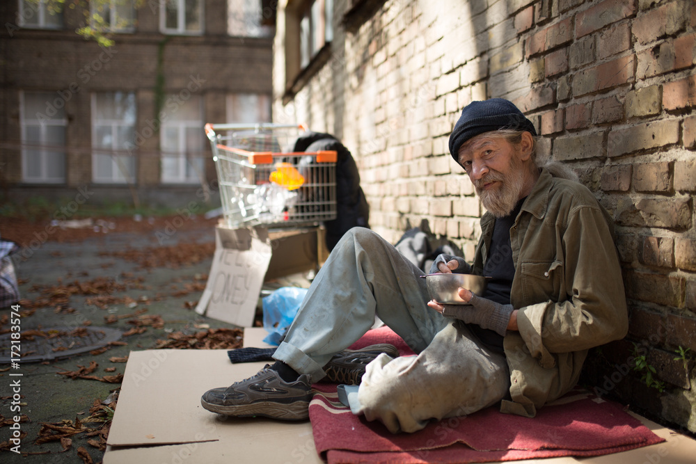 Old homeless man sitting on cardboard holding bowl with food in hand ...