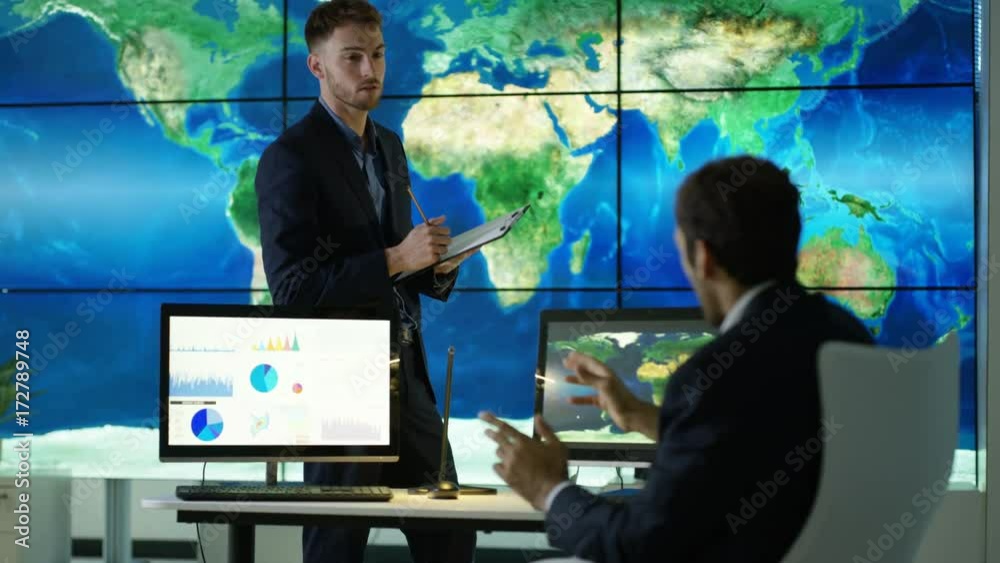 Businessmen discussing large world map graphic on video wall, with pie ...