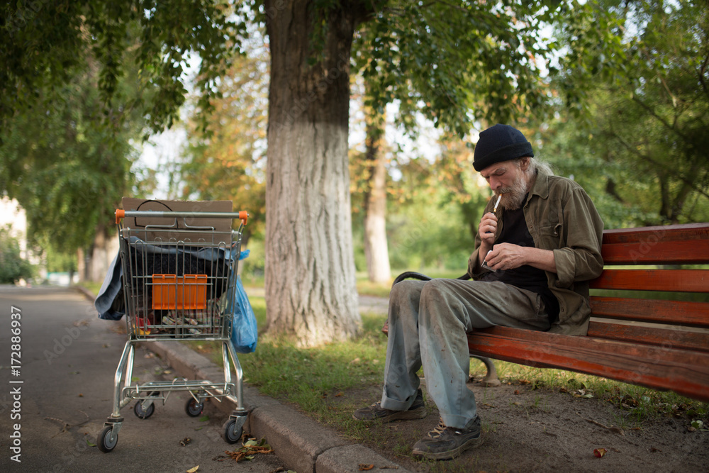 Homeless man sitting on bench lightning cigarette. Old man with no ...
