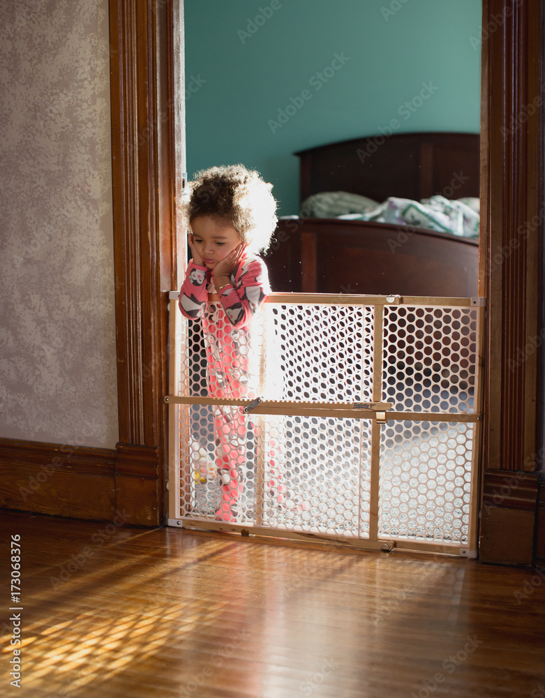 Portrait of toddler girl with curly hair standing behind a safety gate ...