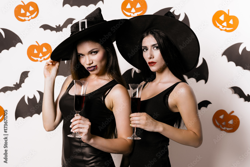 Two sexy witches in black clothes posing against the wall with bats ...
