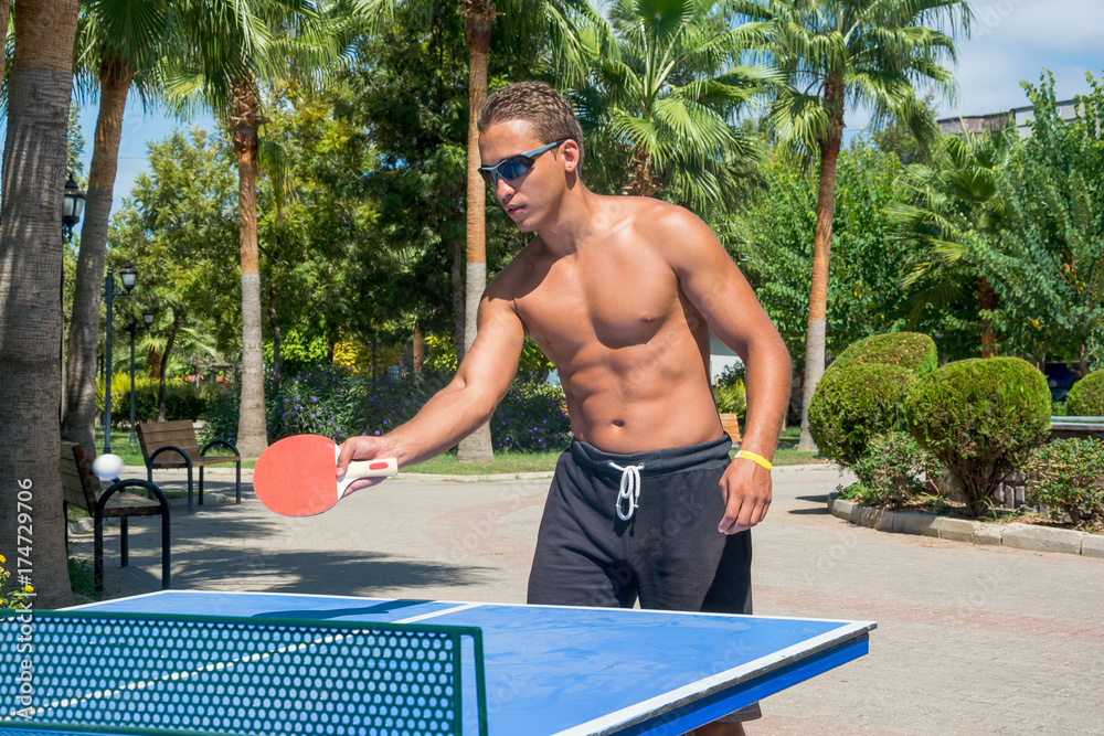 A young guy plays table tennis without shirts in a park on a background ...